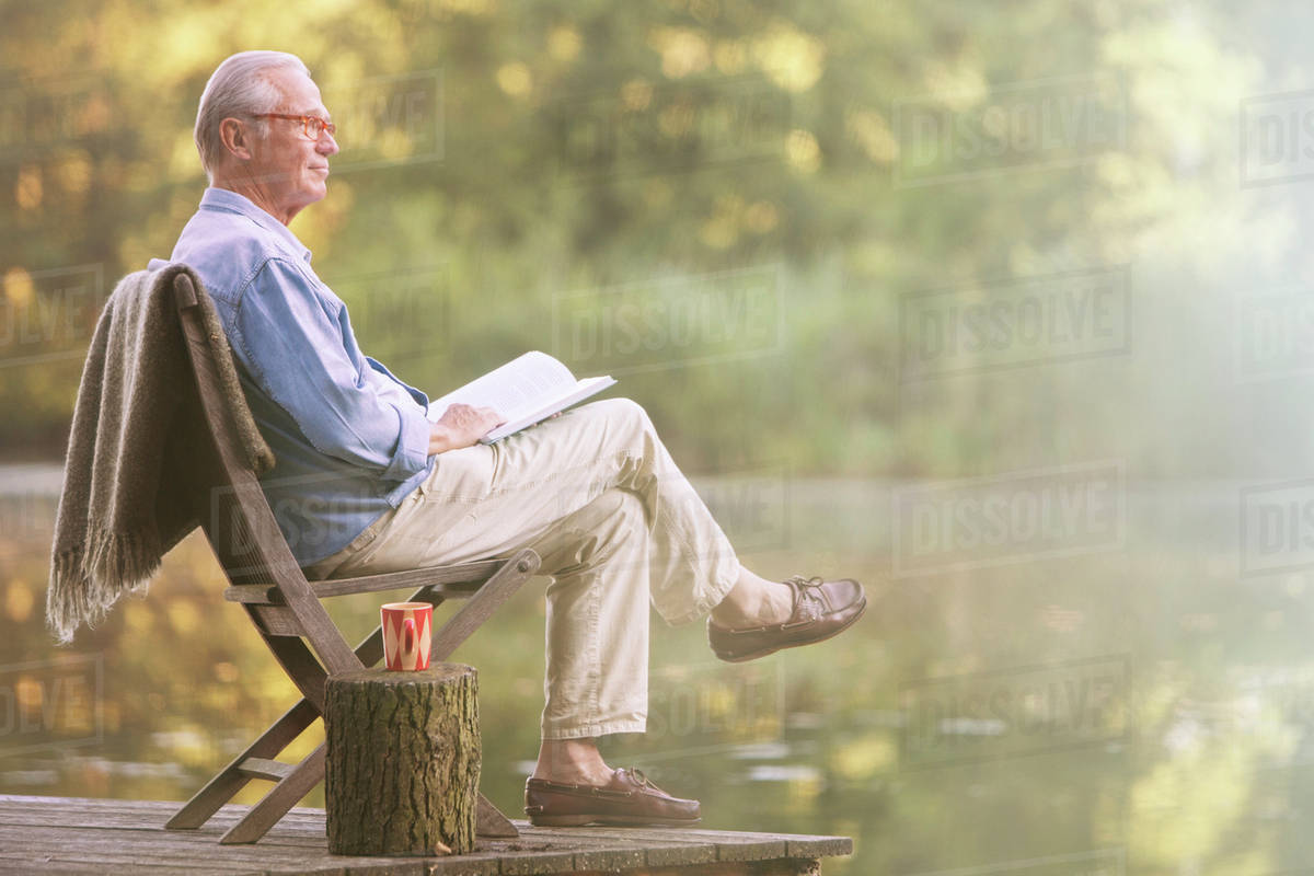 Older man reading book on dock at lake - Stock Photo - Dissolve