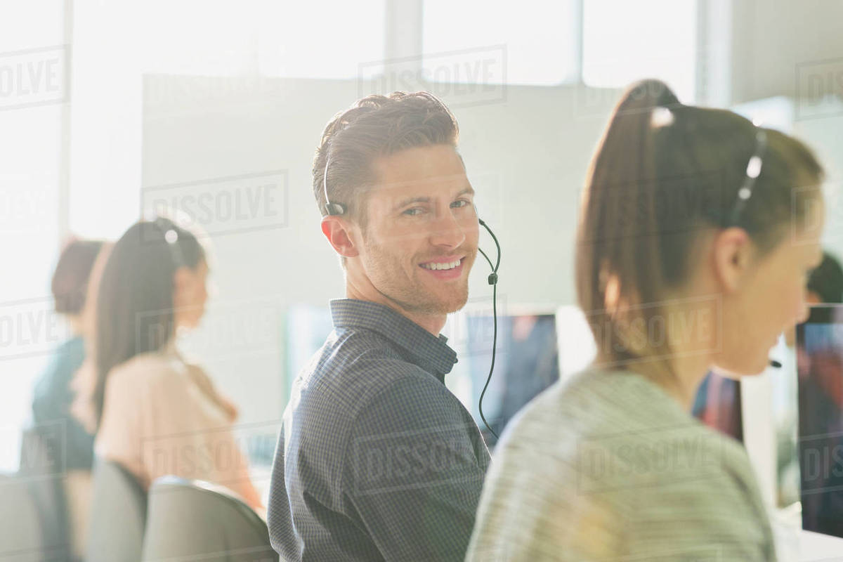 Portrait smiling male telemarketer wearing headset at computer in sunny ...