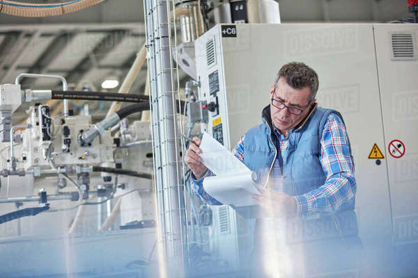 Male supervisor with clipboard talking on cell phone in factory ...