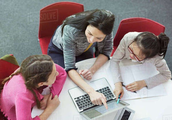 Female teacher helping girl students researching at laptop at library ...