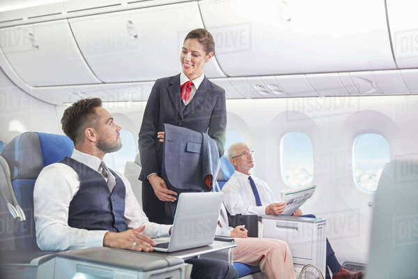 Flight attendant talking with businessman working at laptop on airplane ...