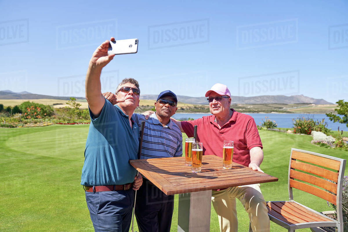 Male friends drinking beer and taking selfie on golf course patio ...