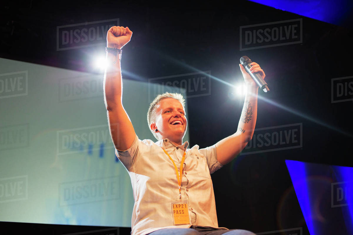 Enthusiastic, smiling female speaker with microphone cheering on stage ...
