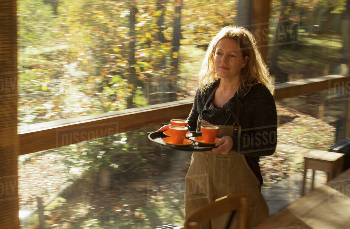Female barista carrying coffee on tray along sunny autumn cafe window ...