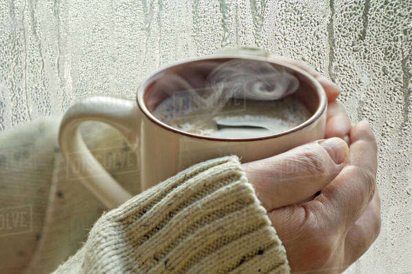 Human hands holding a mug with steam rising up, next to a window with ...