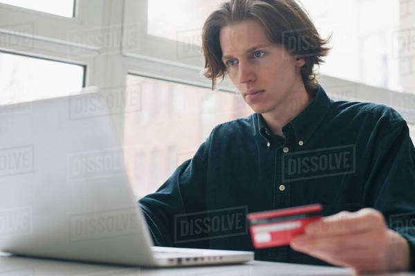 Young man holding credit card while using laptop at table - Royalty ...