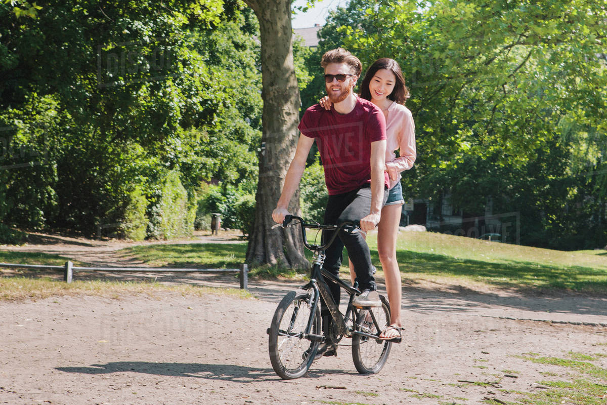 Full length of young couple riding bicycle at park - Stock Photo - Dissolve