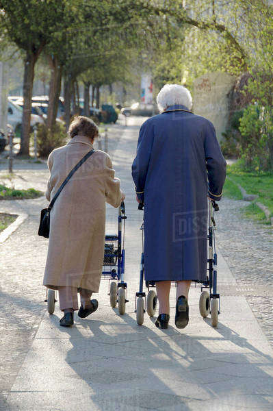 Two women using walkers to walk together on a sidewalk, rear view ...