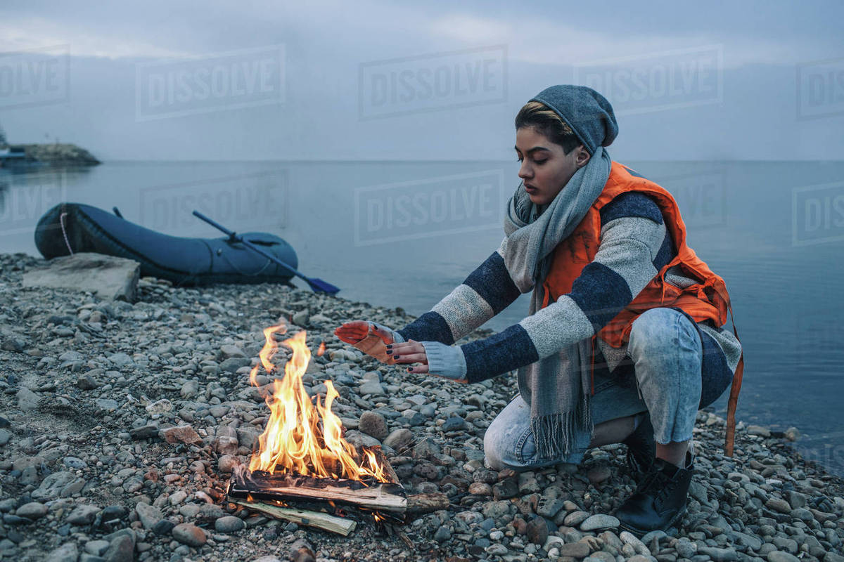 Full length of teenage girl warming hands over bonfire on lakeshore ...