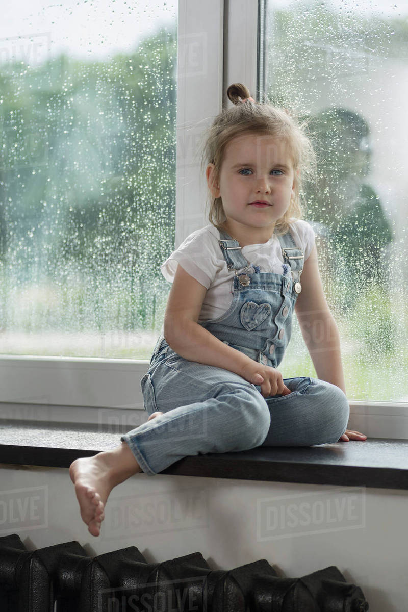 Portrait of cute girl sitting on window sill at home during rainy ...