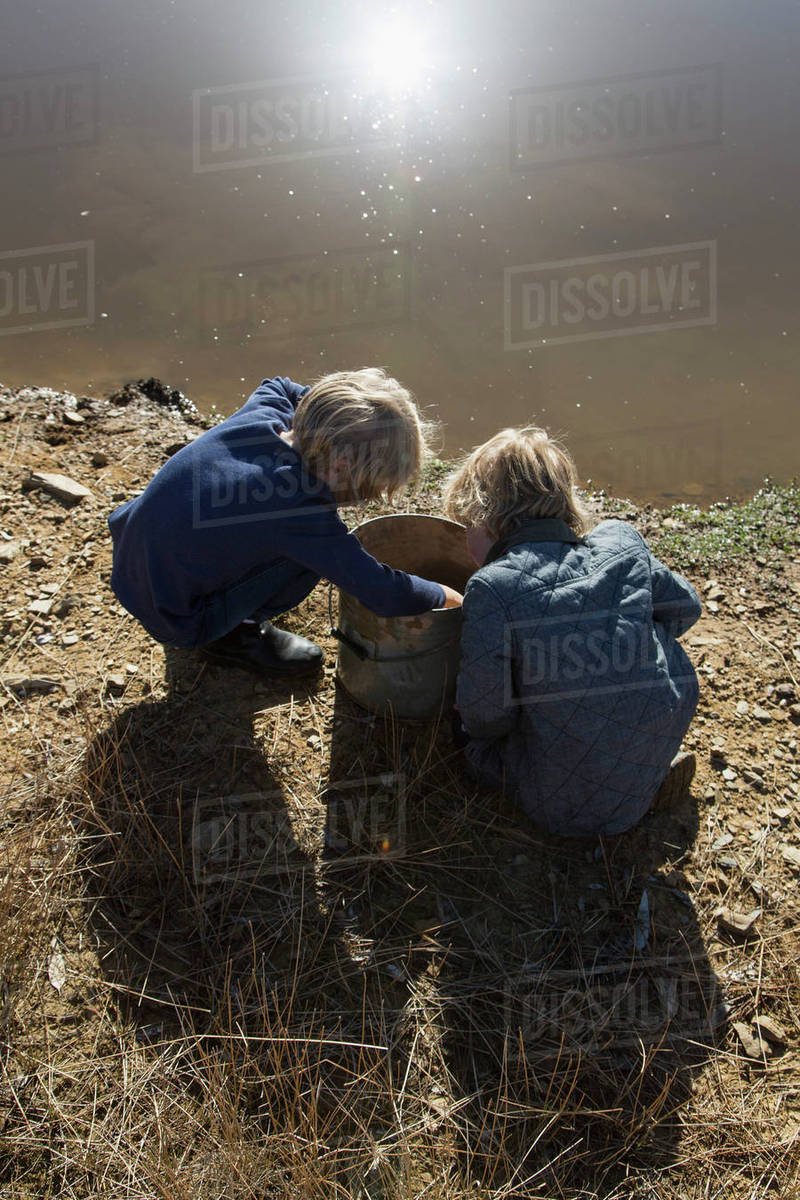 High angle view of boys crouching by bucket at the edge of a river ...