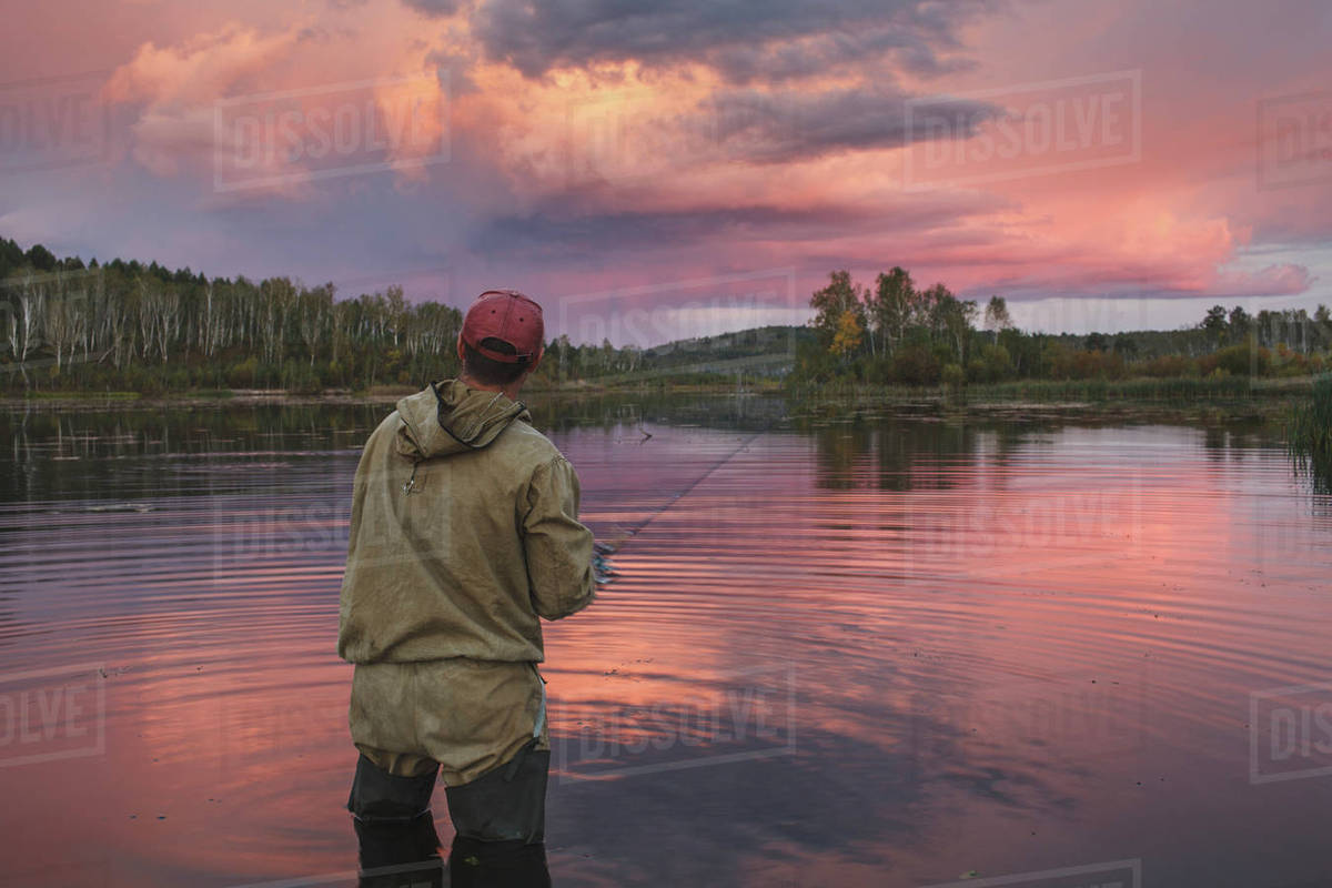Rear view of man fishing in lake against cloudy sky at forest during ...