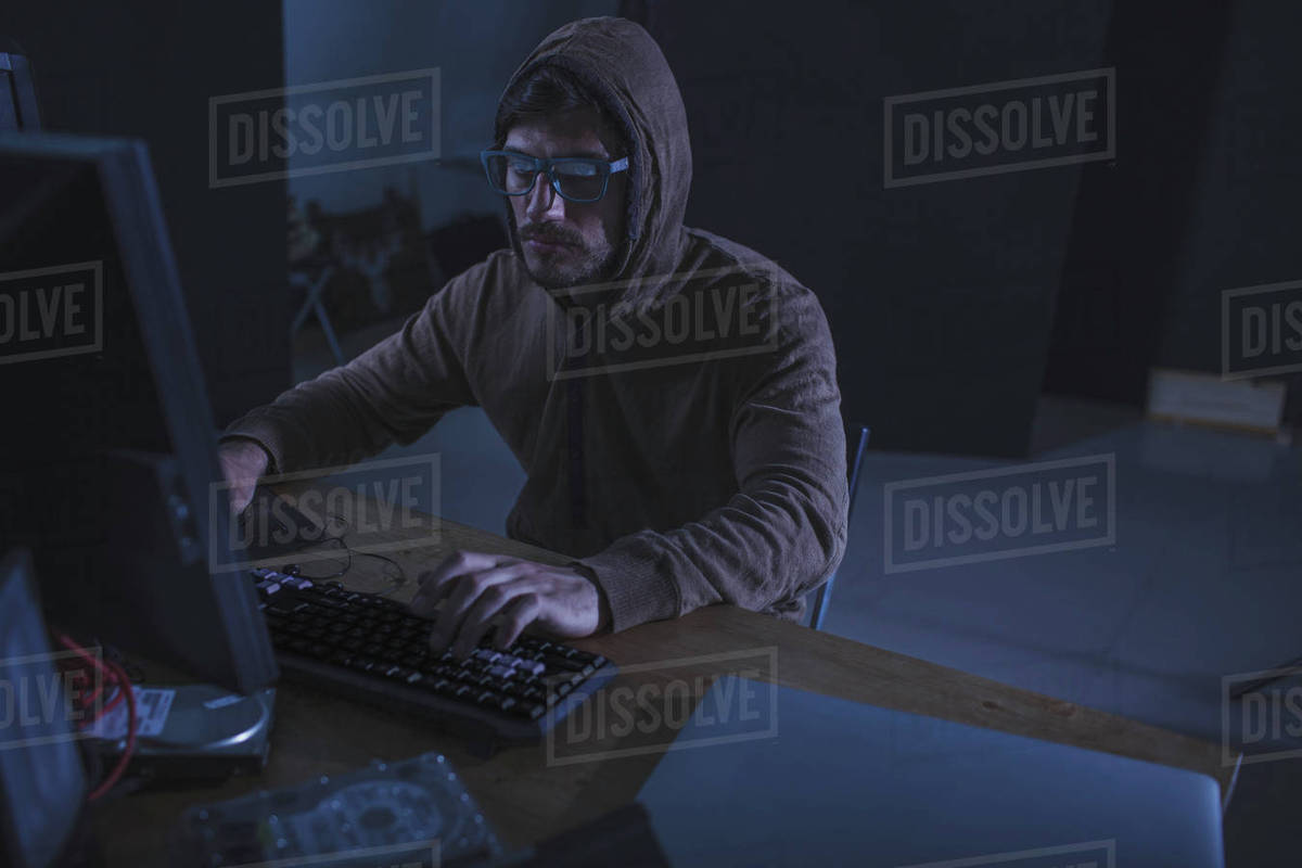 Serious computer hacker wearing hooded shirt using desktop computer at ...