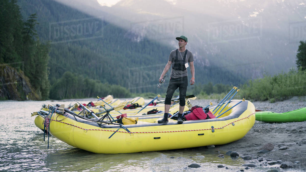Man looking away while standing on raft at riverbank against mountain ...
