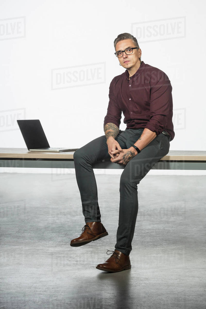 Portrait of man sitting on desk with laptop - Stock Photo - Dissolve